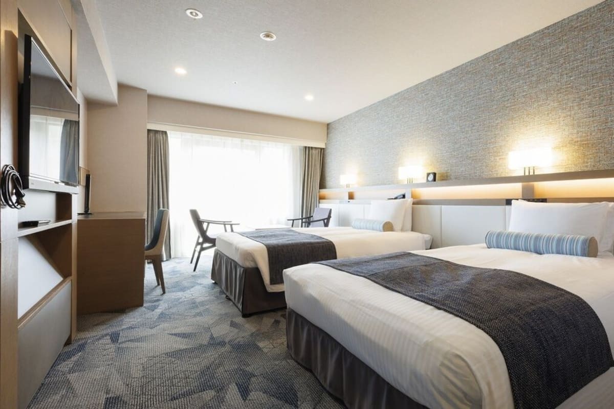 Quarto de hotel com duas camas, uma escrivaninha, uma cadeira e uma televisão.