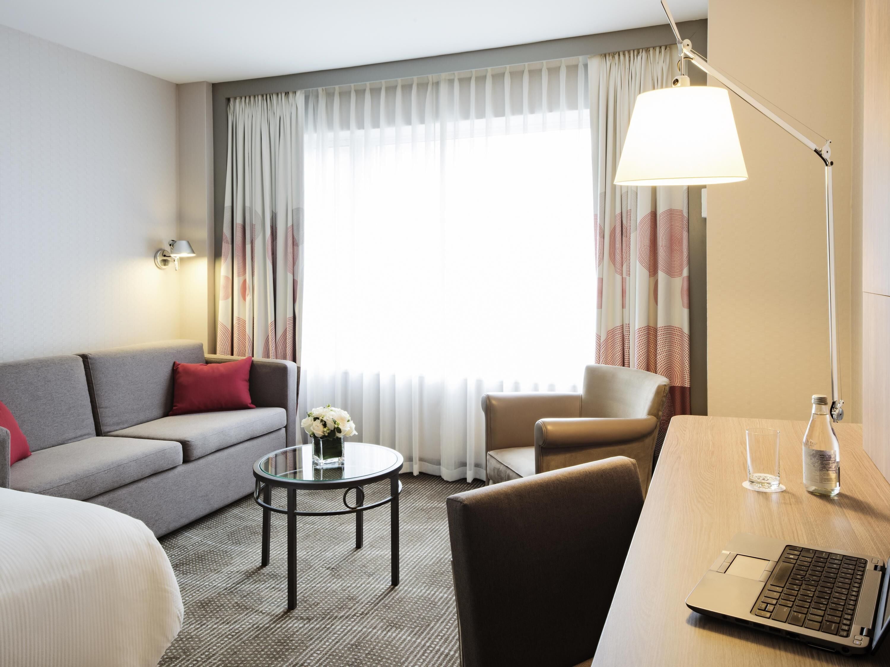 Quarto de hotel com sofá, poltrona, mesa de centro redonda e escrivaninha com um laptop.