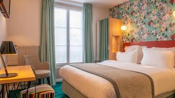 Quarto de hotel com cama, escrivaninha, cadeira, radiador e janela com cortinas.