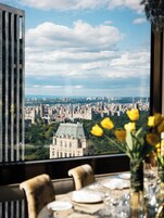 Uma sala de jantar com uma mesa posta para dois, um vaso com flores amarelas e vista para a paisagem urbana, com um edifício em destaque ao primeiro plano.