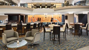 Um saguão de hotel moderno, com um bar central, diversas áreas de estar e teto alto com iluminação embutida.