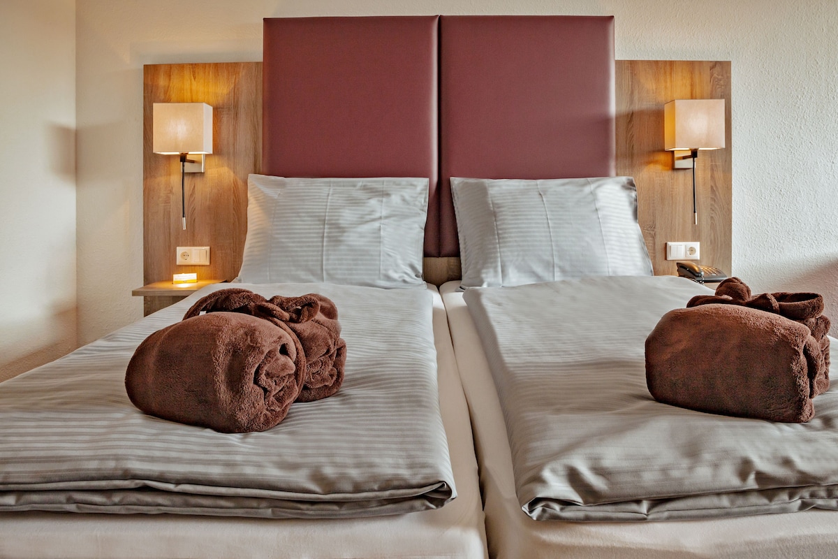 Quarto de hotel com duas camas, cada uma com uma toalha castanha e uma mesa de cabeceira com abajur.