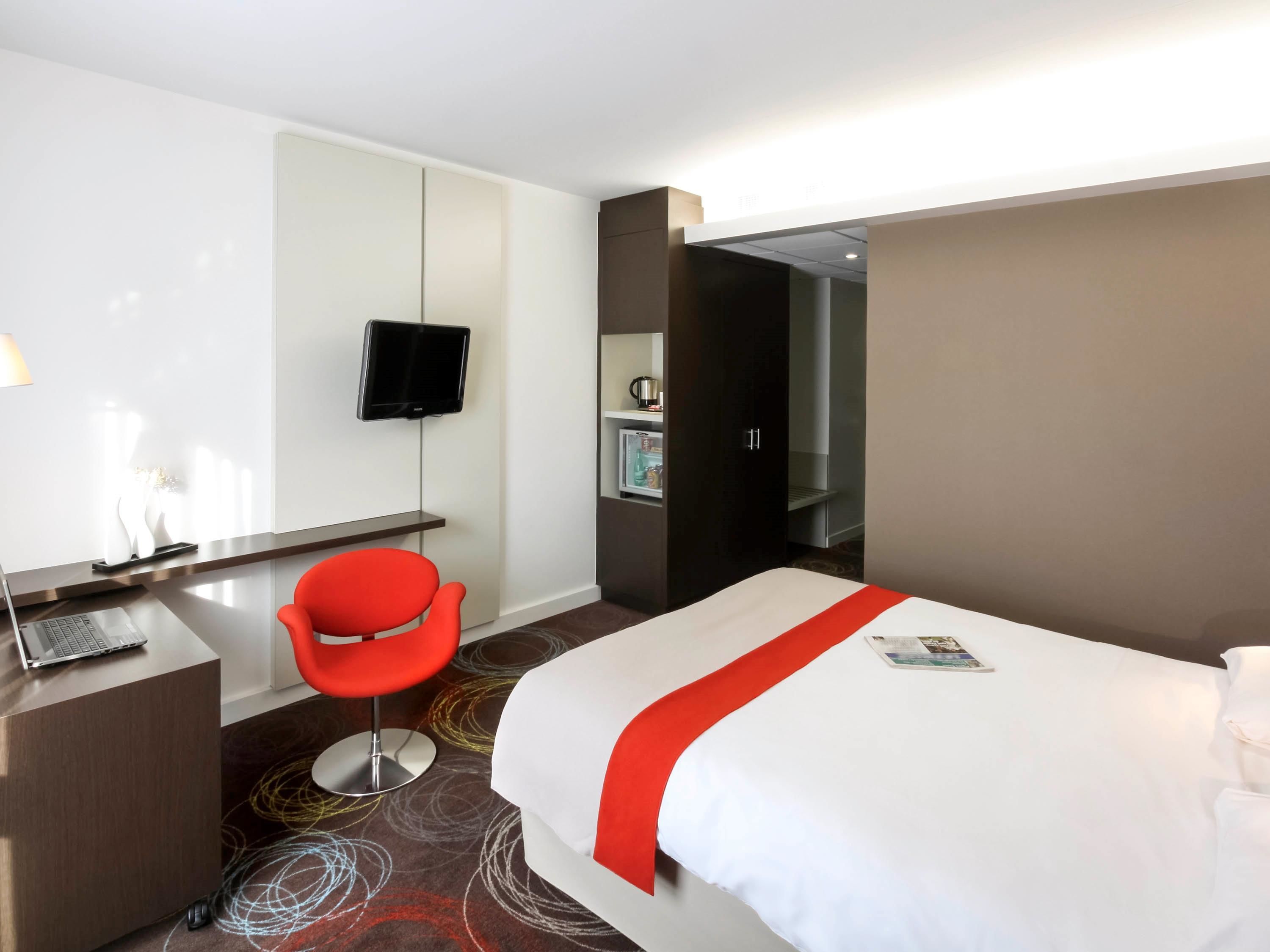 Quarto de hotel com cama, escrivaninha, cadeira, televisão e minibar.