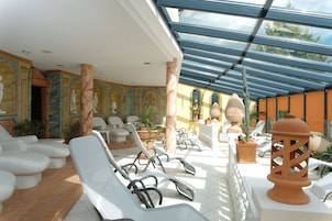 Pátio interno iluminado pelo sol, com cadeiras de lounge brancas, teto de vidro e colunas decorativas.