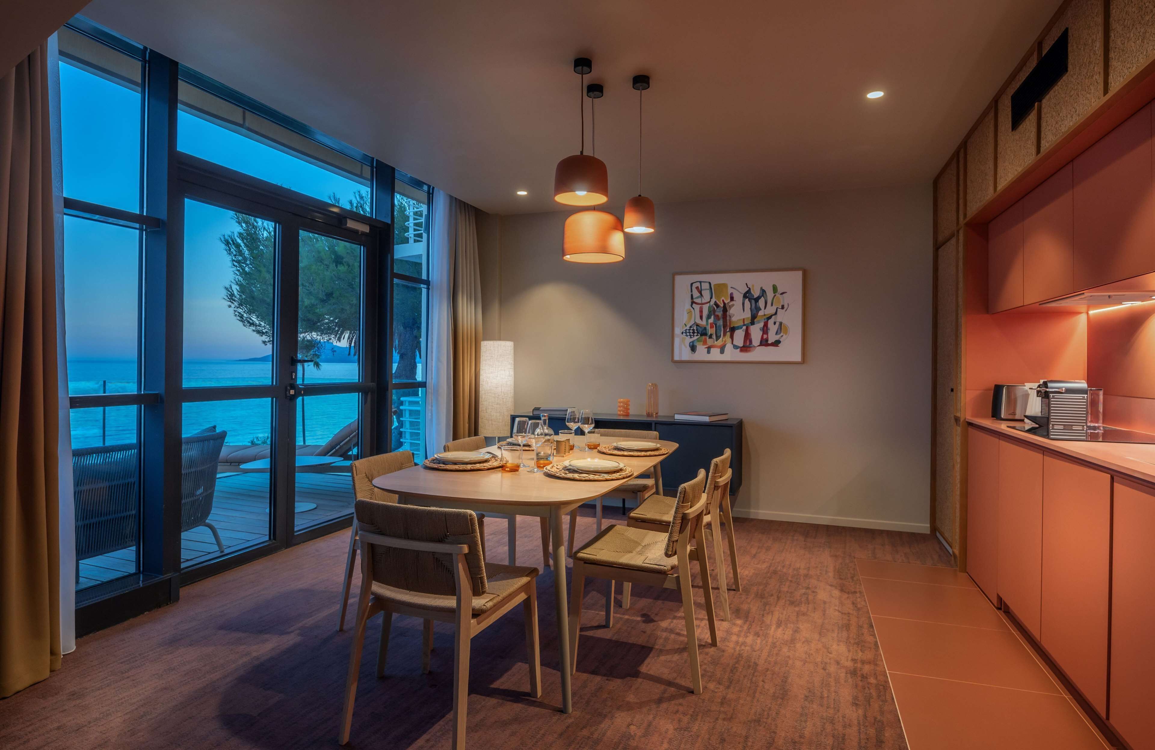 Uma área de refeições com vista para o mar, uma mesa redonda posta para quatro pessoas e uma cozinha com armários e bancada.