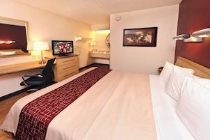 Quarto de hotel com cama, escrivaninha com computador, cadeira, espelho, abajur e quadro pendurado na parede.