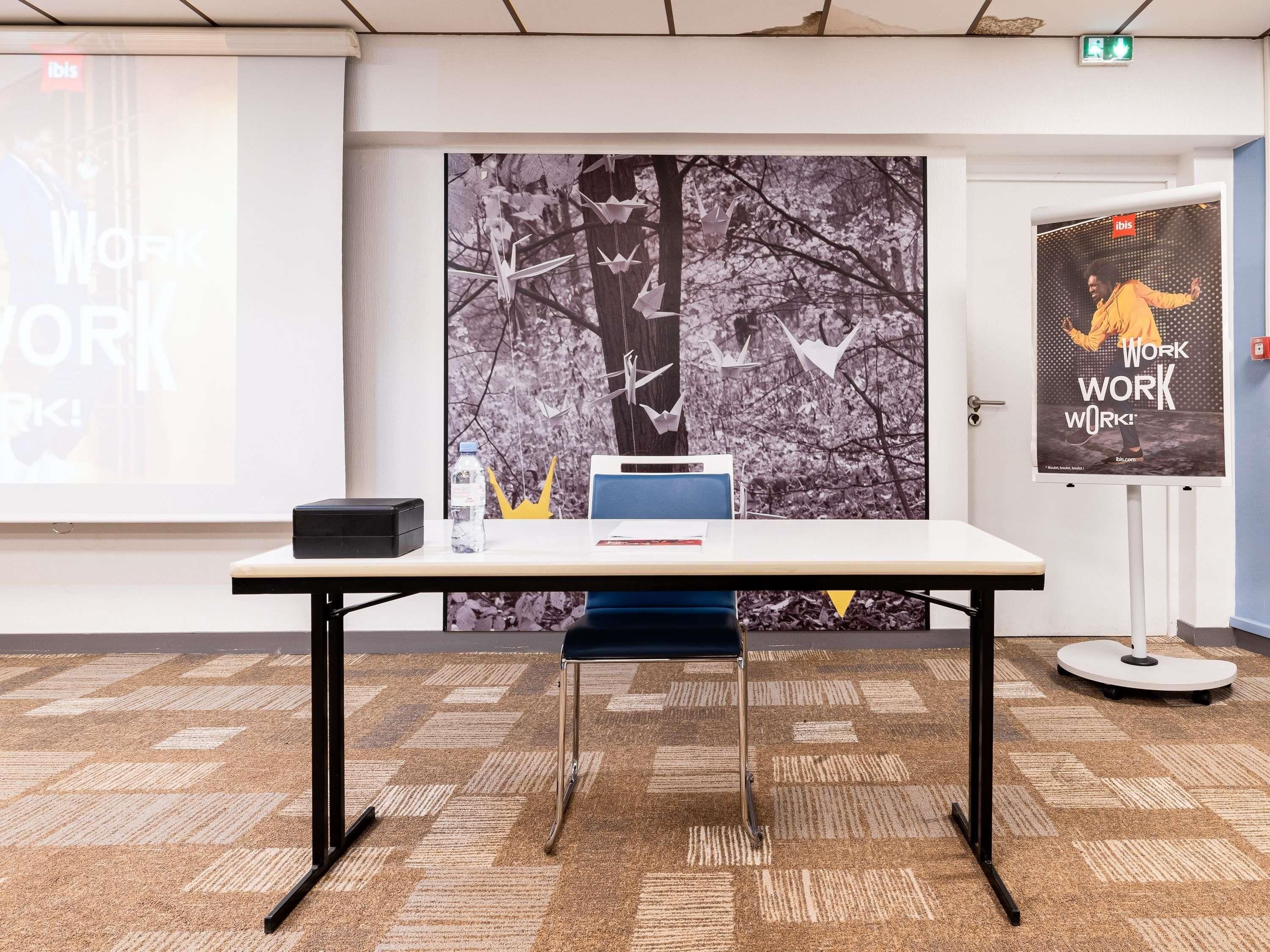 Uma sala de conferência com tela de projetor, uma mesa com um laptop, uma garrafa de água e uma cadeira.