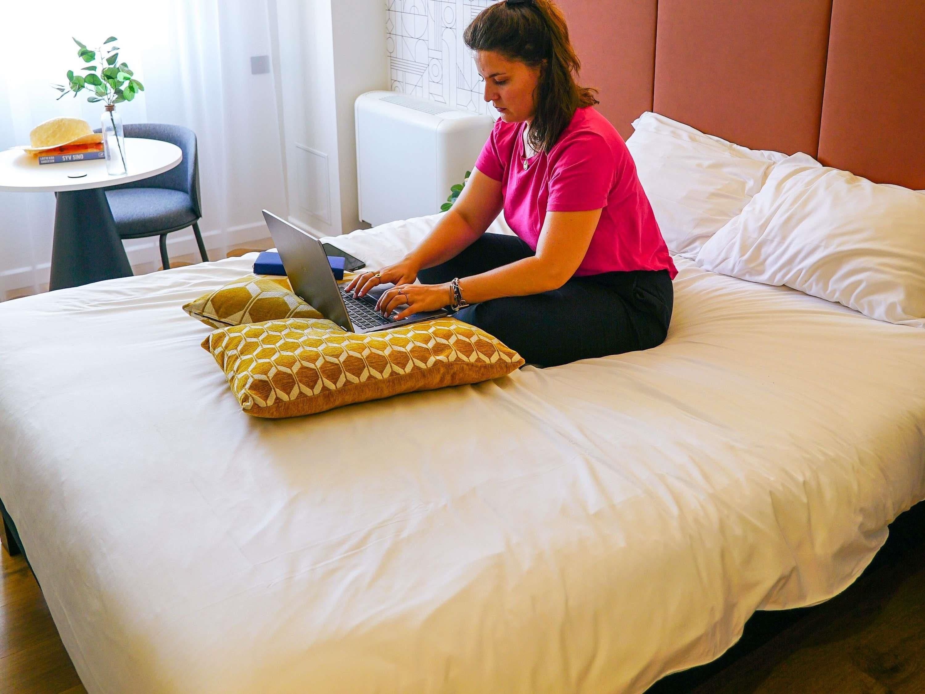 Uma pessoa sentada em uma cama com um laptop, almofadas, uma mesinha com uma planta e uma cadeira.
