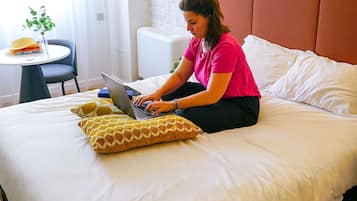 Uma pessoa sentada em uma cama com um laptop, almofadas, uma mesinha com uma planta e uma cadeira.