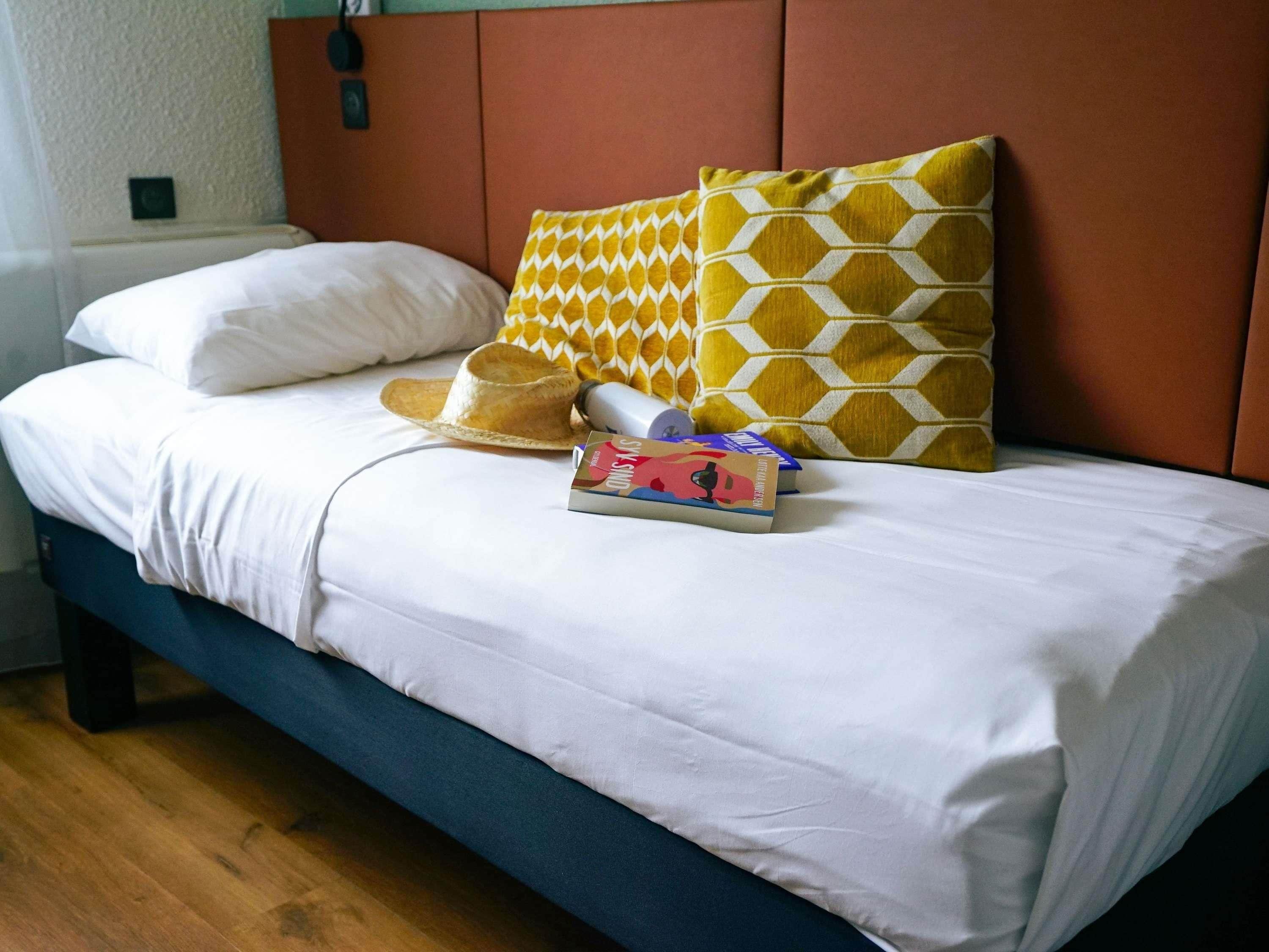 Cama bem arrumada com roupa de cama branca, cabeceira marrom e travesseiro com padrão amarelo.