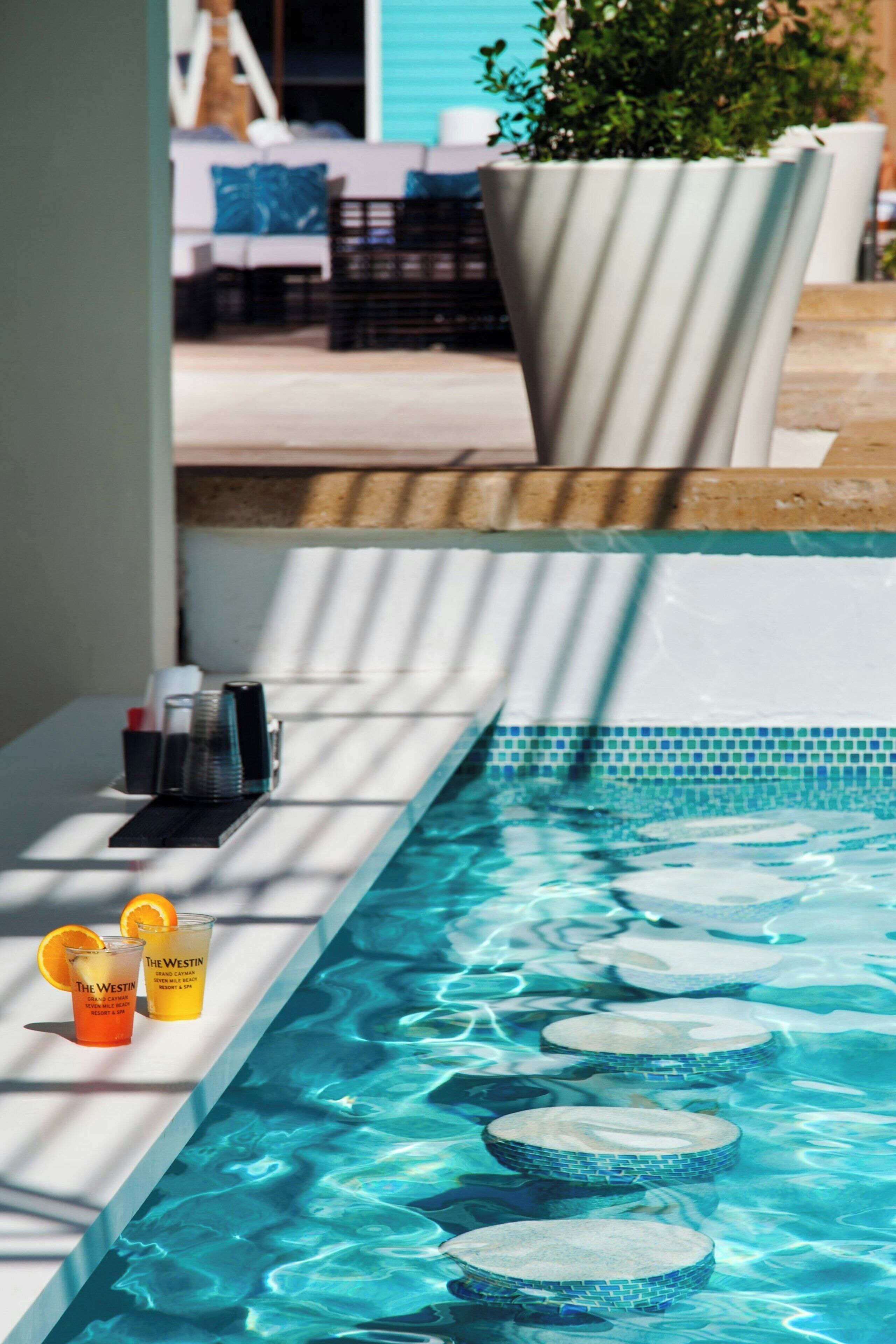Área da piscina com disposição de assentos, um vaso de planta e dois copos da bebida Westin.