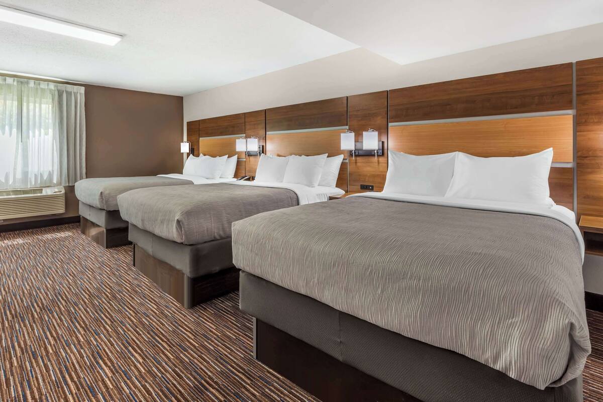 Quarto de hotel com duas camas, uma janela grande com cortinas, cabeceira de madeira e carpete estampado.