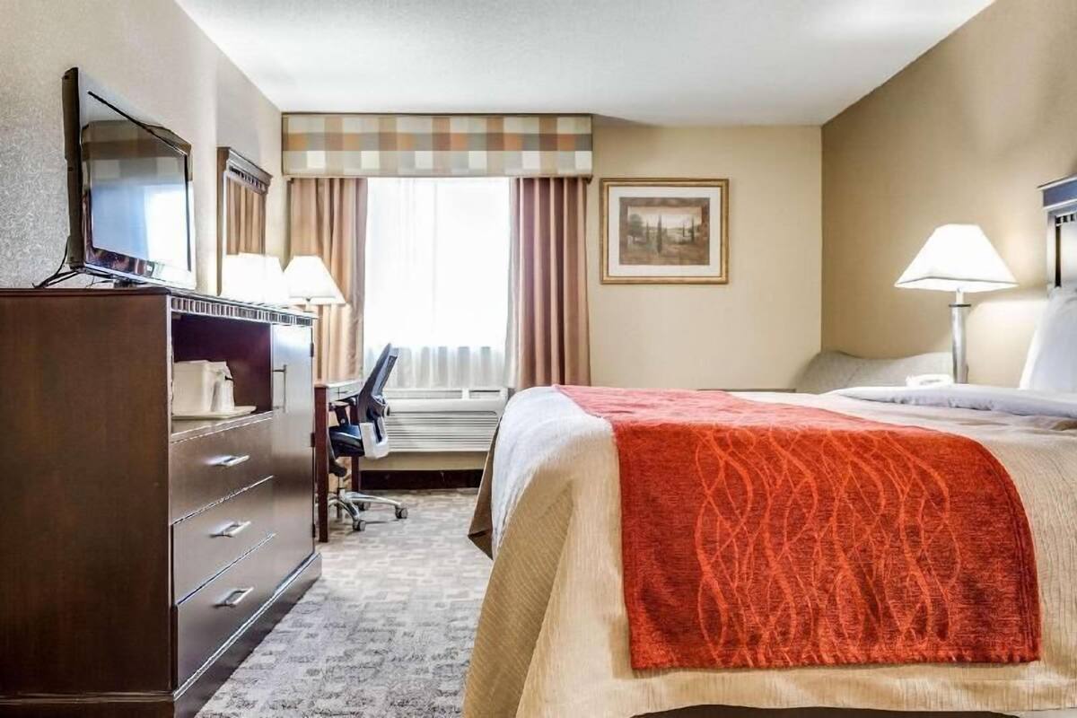 Quarto de hotel com cama, escrivaninha, cadeira, televisão e janela com cortinas.