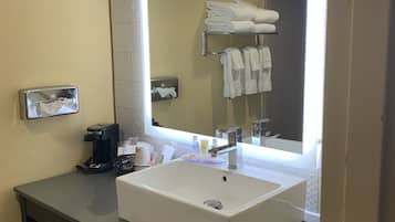Casa de banho de hotel com um espelho grande, uma sanita, uma máquina de café, um dispensador de papel higiénico e uma toalheira com toalhas dobradas.