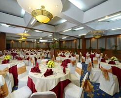 Salão de banquetes com mesas redondas, toalhas brancas e cadeiras adornadas com fitas vermelhas e douradas.