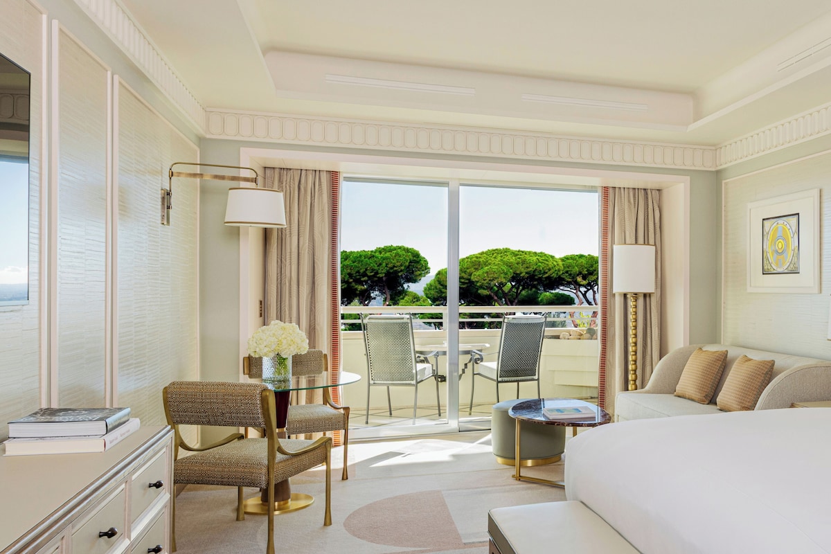 Quarto de hotel com cama, escrivaninha com luminária, cadeira, mesinha e vista para o exterior.