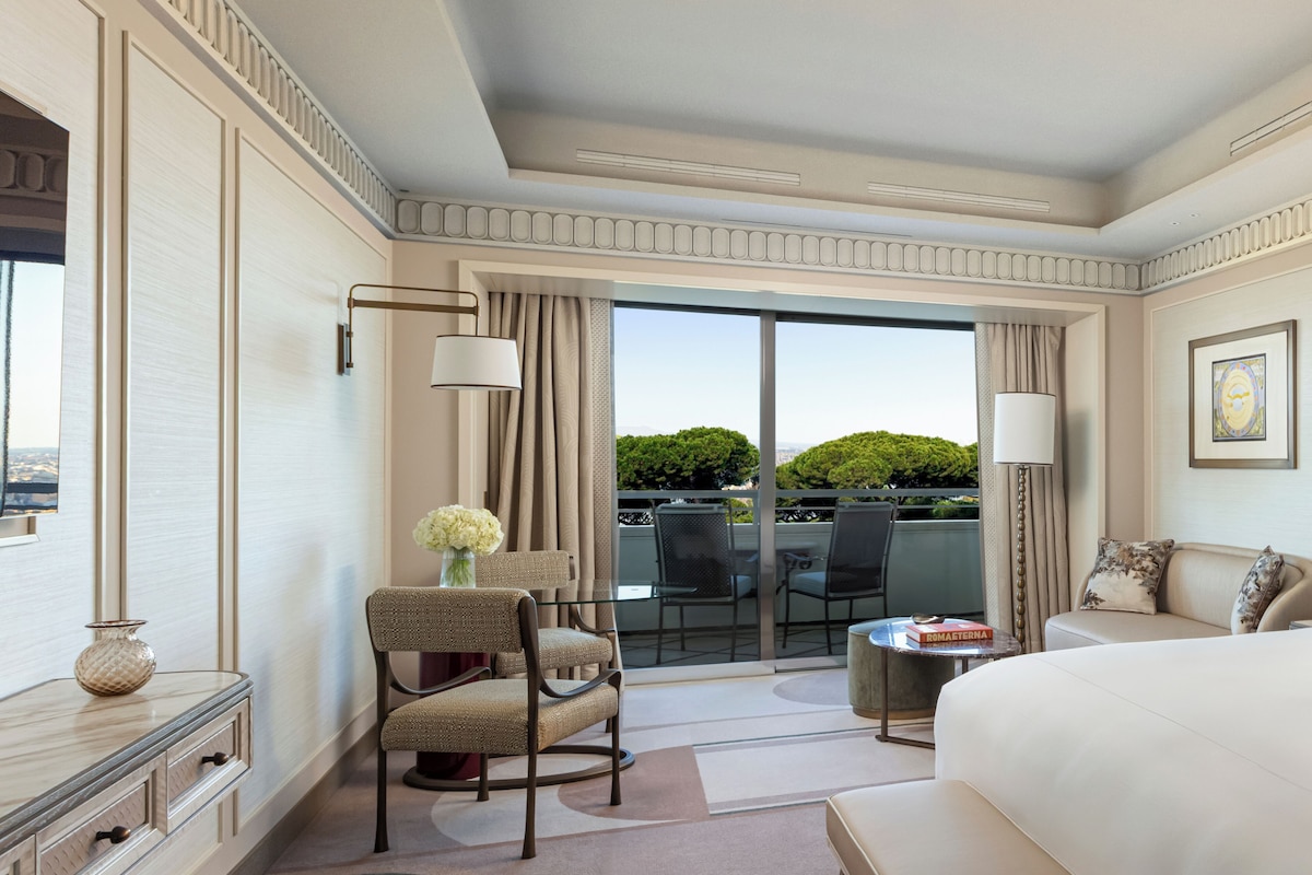 Quarto de hotel com cama, área de estar com cadeiras, mesa e uma grande janela com vista para o verde.