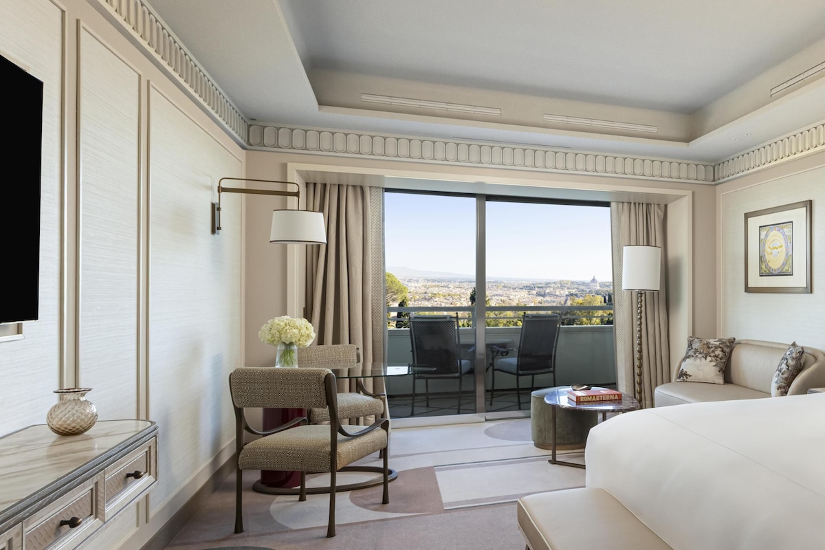 Quarto de hotel com uma cama grande, uma escrivaninha, uma cadeira, uma televisão, uma varanda com vista e um vaso com flores.