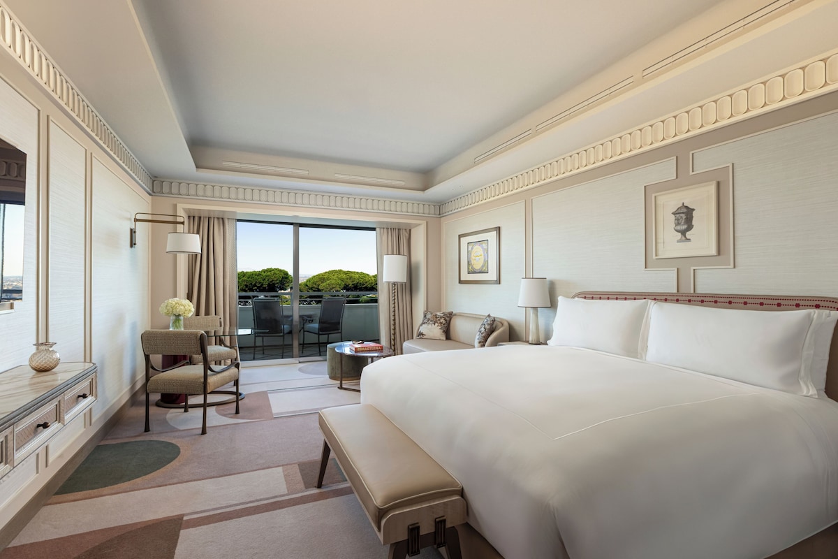Quarto de hotel com uma cama grande, escrivaninha, cadeira, mesa pequena e vista para o exterior.