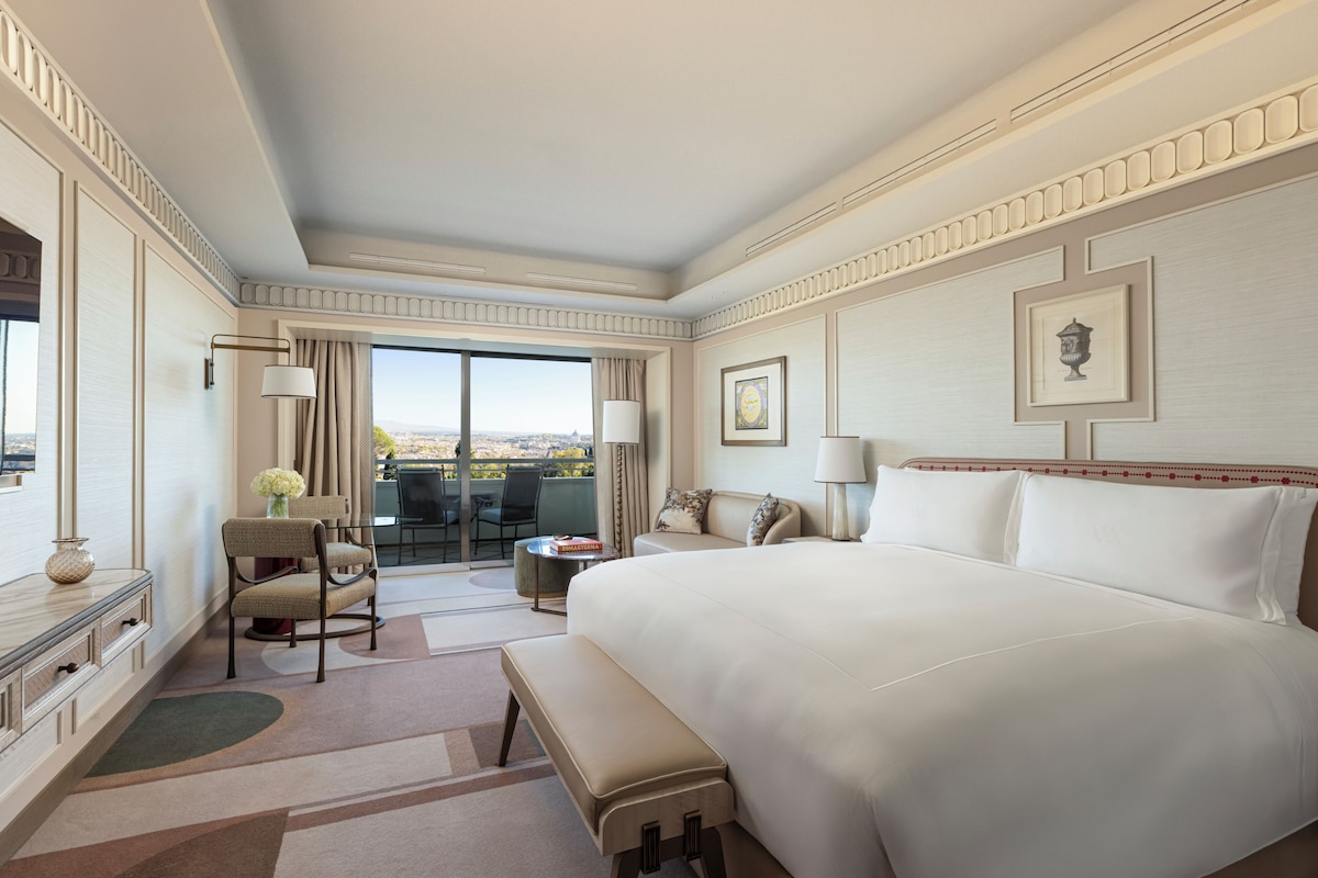 Quarto de hotel com uma cama grande, uma escrivaninha, uma cadeira, uma mesinha e varanda com vista.