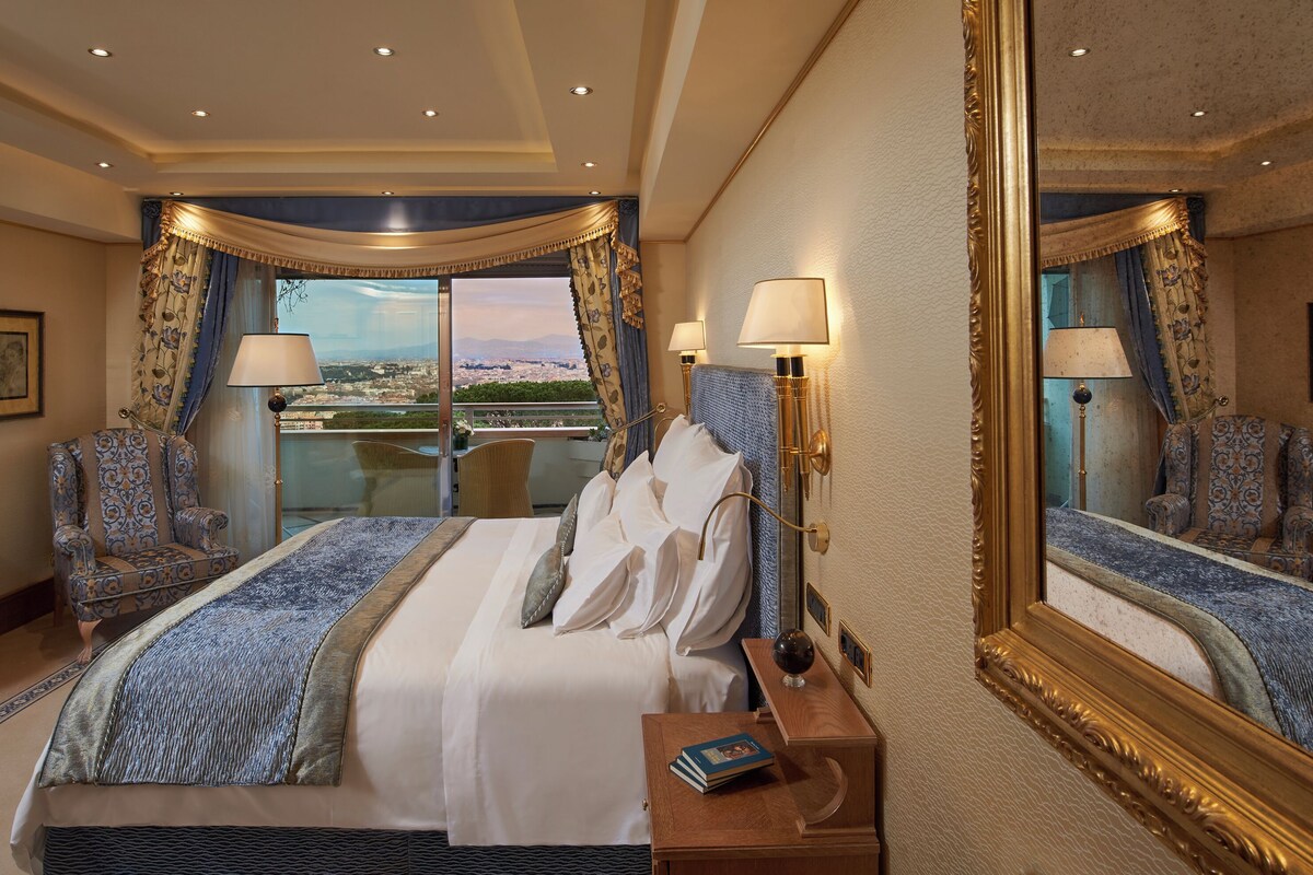Quarto de hotel com uma cama grande, um espaço de estar com uma poltrona, uma escrivaninha e um espelho amplo.