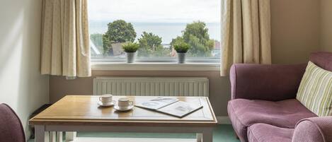 Quarto com vista para o mar, janela com cortinas, mesa com xícara e livro, e sofá roxo.