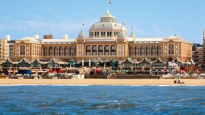 Um edifício grande e histórico com uma cúpula imponente e várias bandeiras, localizado à beira de uma praia com vista para o oceano.