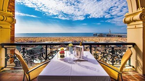 Uma varanda com mesa posta para dois, com vista para uma praia e um cais ao longe.