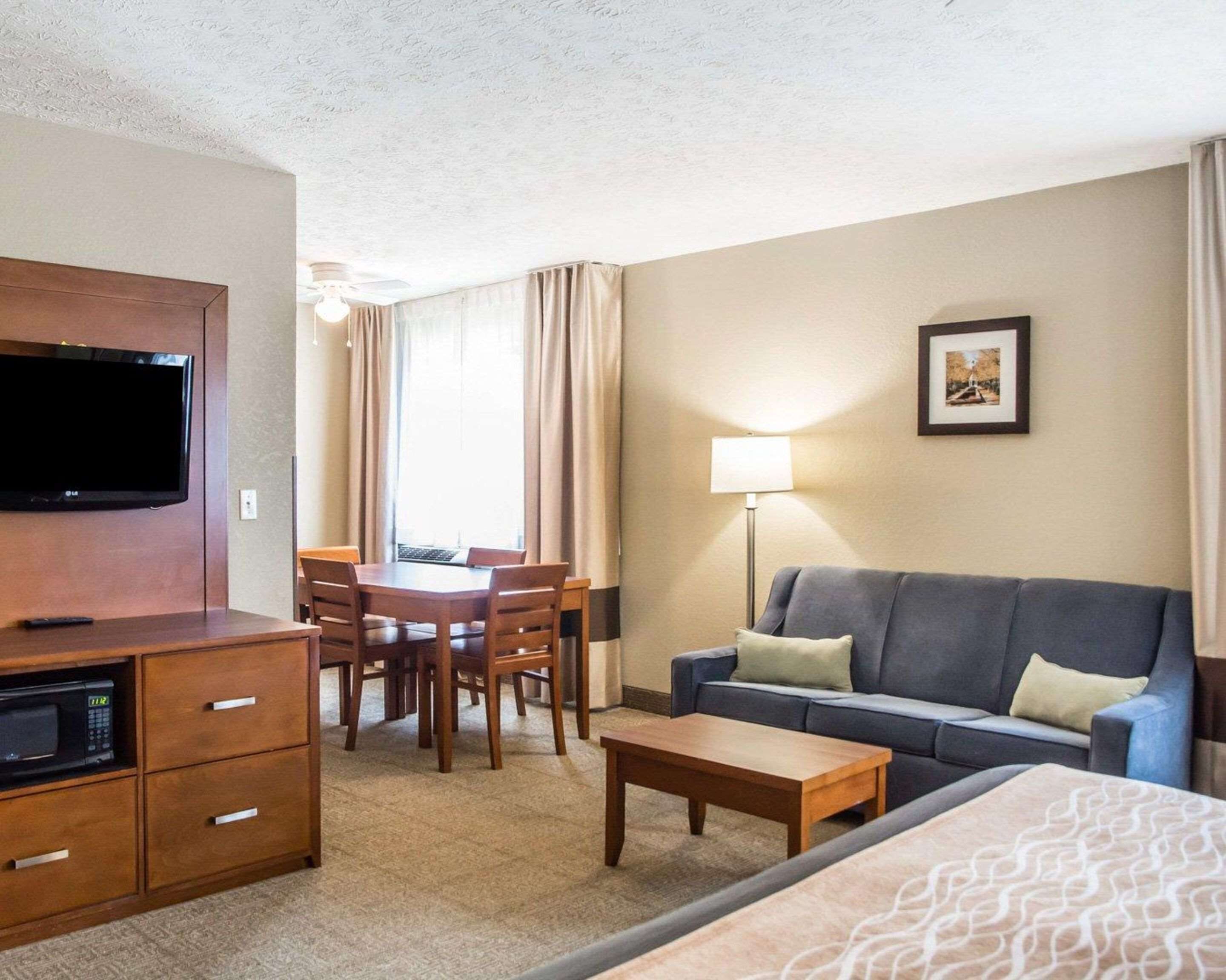 Quarto de hotel com televisão de tela plana, mesa de jantar com cadeiras, sofá, luminária e um quadro na parede.