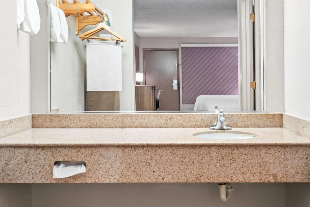 Casa de banho de hotel com lavatório de bancada em granito, um porta-toalhas com toalhas brancas e um espelho refletindo um quarto com uma cama e uma parede listrada de roxo.