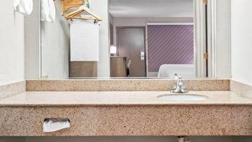 Casa de banho de hotel com lavatório de bancada em granito, um porta-toalhas com toalhas brancas e um espelho refletindo um quarto com uma cama e uma parede listrada de roxo.