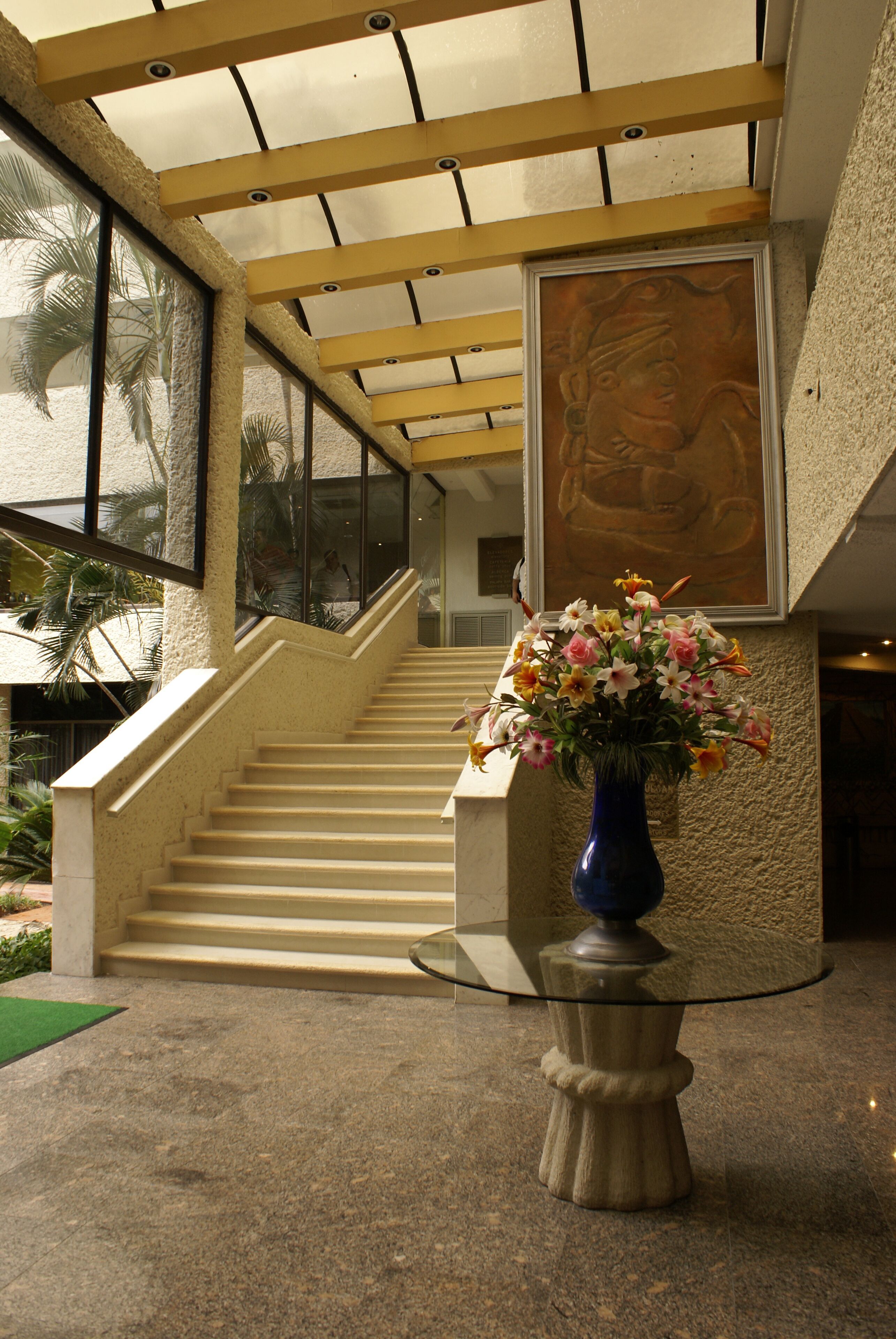 Um hall de hotel com uma mesa de vidro que suporta um vaso de flores, uma escadaria e um grande mural na parede.