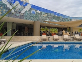 Uma área de piscina com espreguiçadeiras e plantas em vasos, um mural com animais selvagens e uma piscina azul cristalina.
