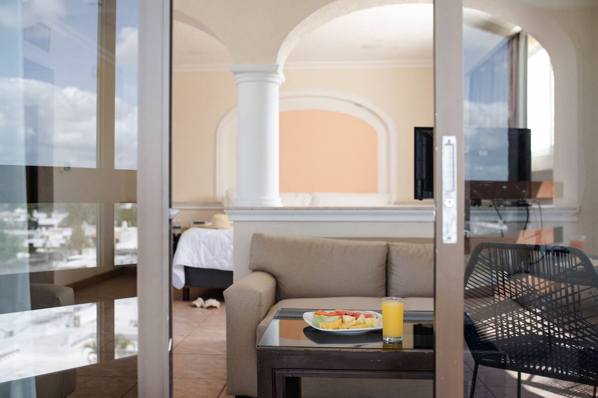 Uma varanda com porta de vidro, um sofá, uma mesa com um prato de comida e um copo de suco de laranja, e uma cadeira preta.