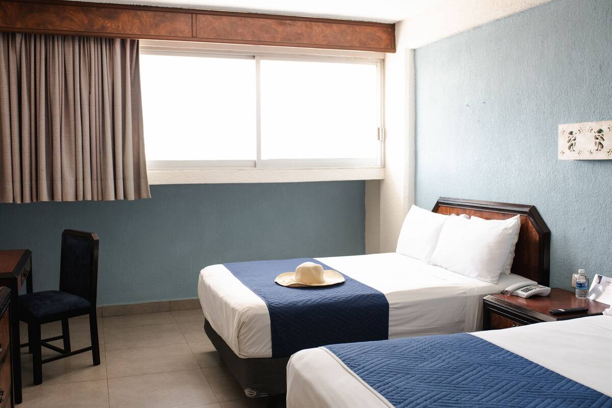 Quarto de hotel com duas camas, uma escrivaninha, uma cadeira, janela com cortinas e mesa de cabeceira com telefone e garrafa.