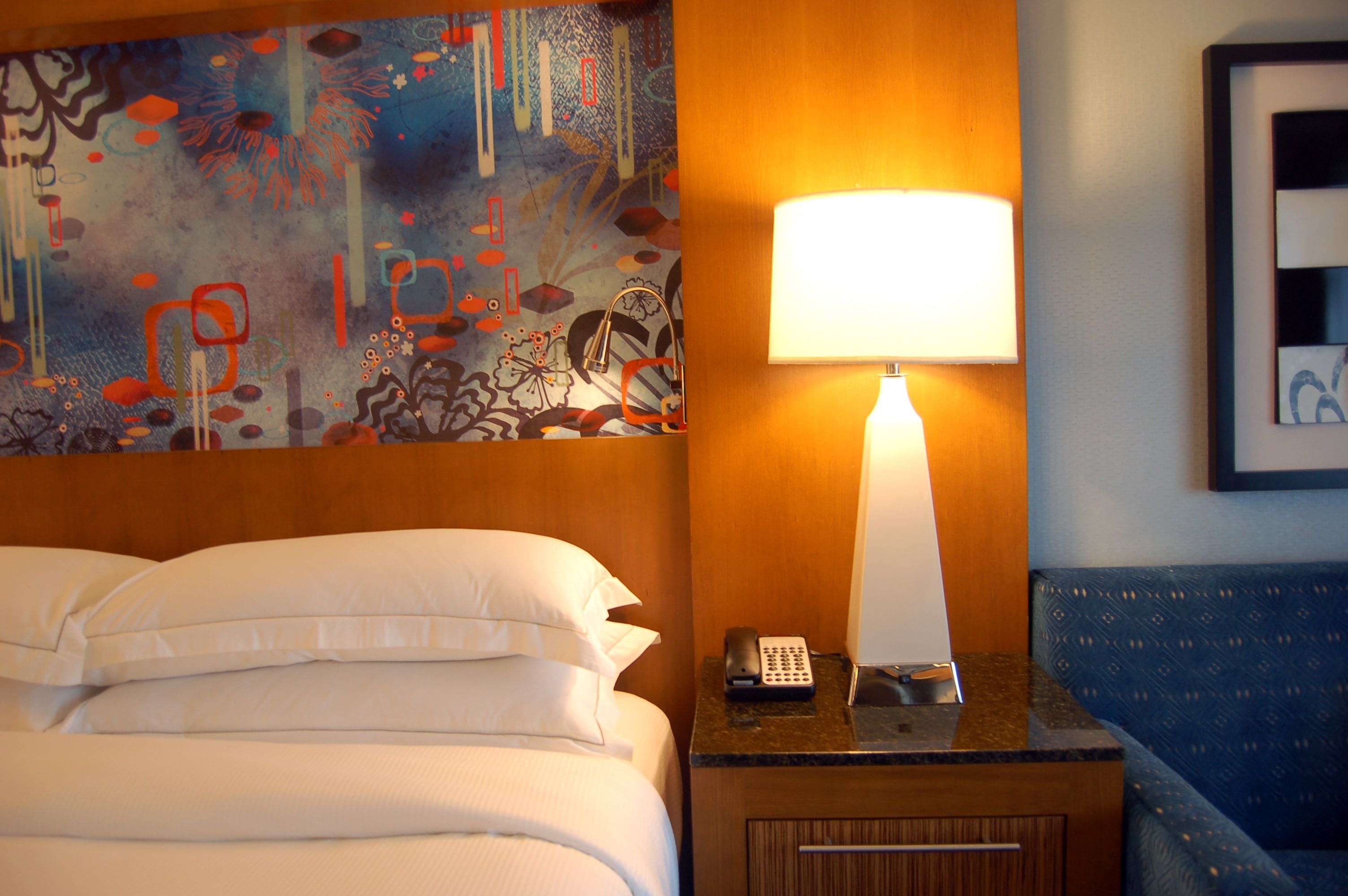 Quarto de hotel com cama, mesa de cabeceira com luminária, telefone e uma obra de arte na parede.