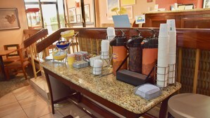 Um balcão de café da manhã com máquinas de café, uma cesta de frutas e empilhamento de copos de papel.