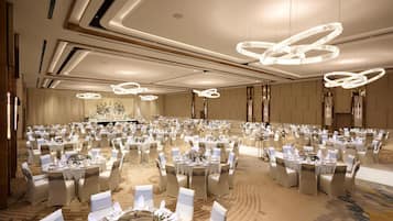Um amplo salão de festas com mesas redondas preparadas para eventos, com luminárias modernas e piso coberto por carpete.