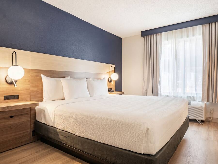 Quarto de hotel com uma cama grande, cabeceira de madeira, luminárias de cabeceira, mesa de apoio, janela com cortinas e um aparelho de ar condicionado.
