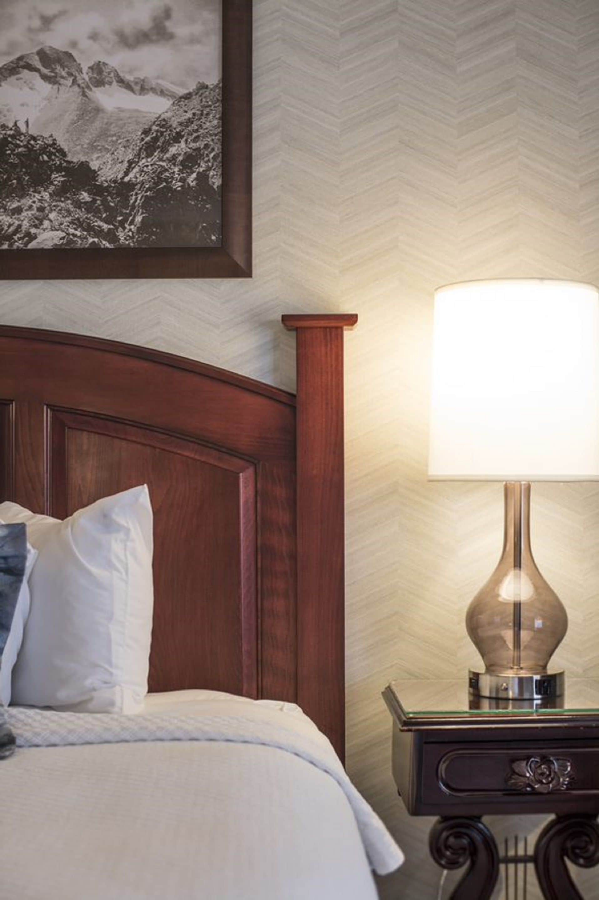 Quarto de hotel com cabeceira de madeira, cama com roupa de cama branca, mesa de cabeceira com abajur e quadro emoldurado na parede.