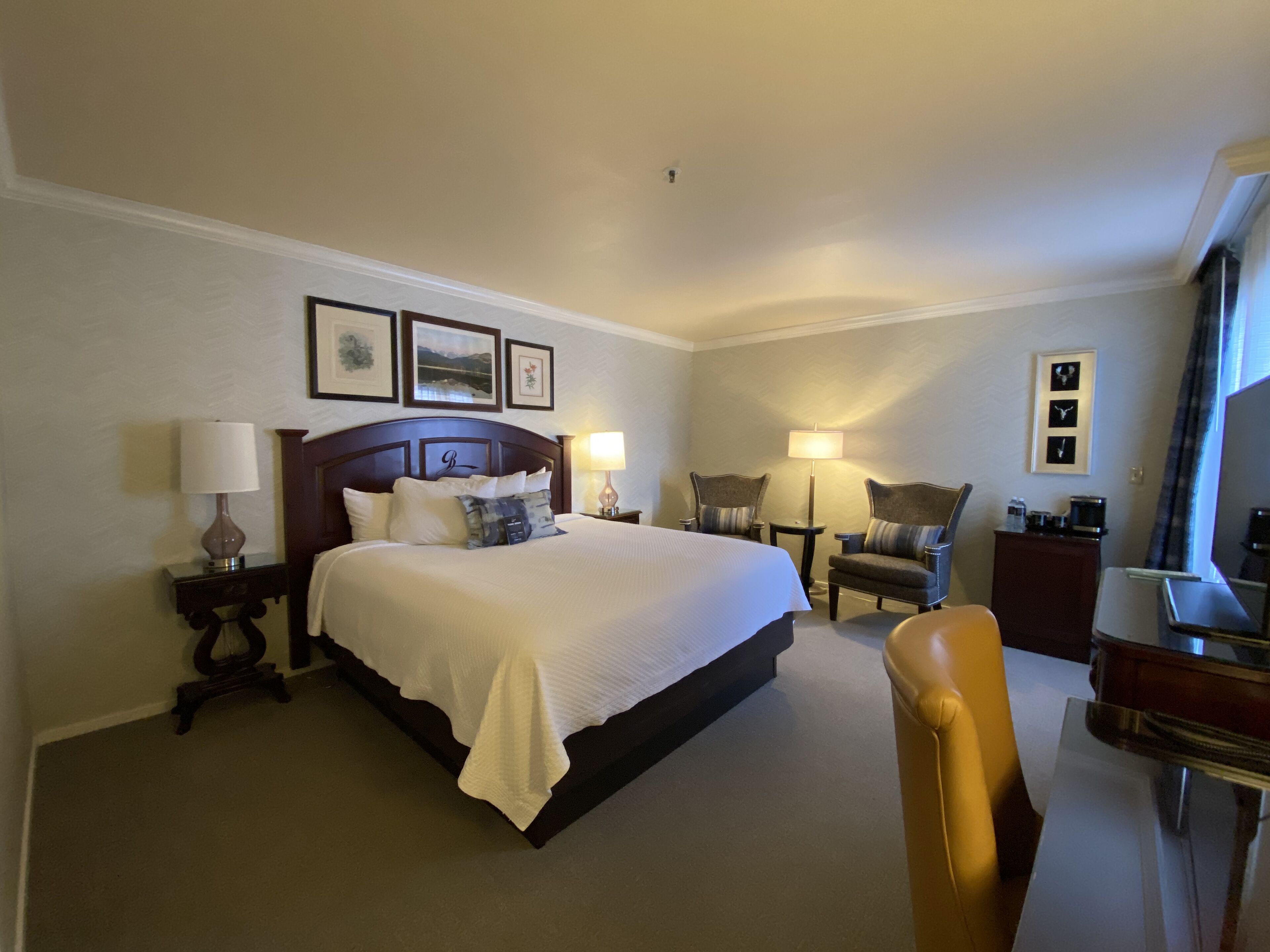 Quarto de hotel com uma cama grande, duas poltronas, uma escrivaninha e uma televisão.