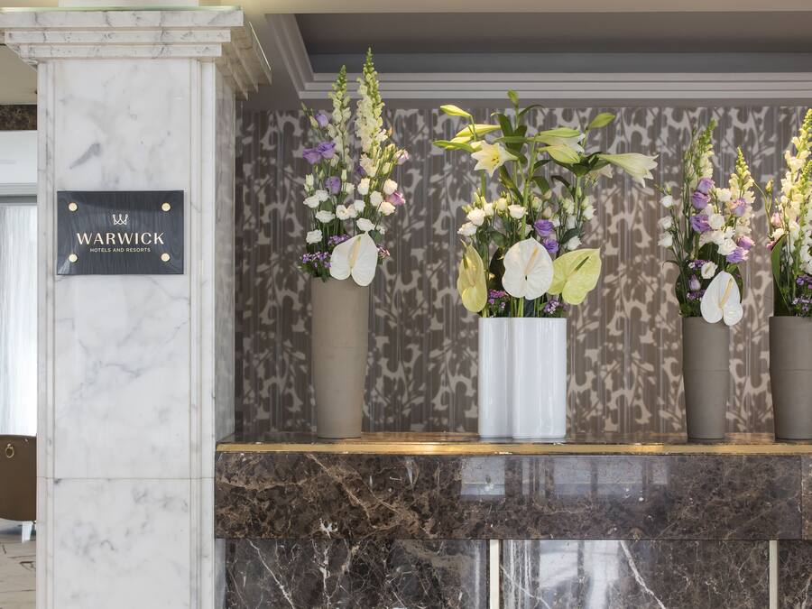 Uma área de recepção com um balcão de mármore, um letreiro indicando "Warwick Hotels and Resorts" e três vasos de flores.