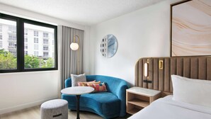 Quarto de hotel moderno com sofá azul curvo, mesa redonda, cama com cabeceira e janela ampla com vista para edifícios e vegetação.