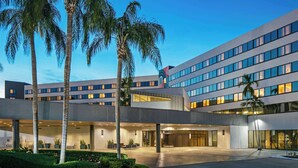 Um hotel moderno com estrutura em vários andares, entrada coberta e palmeiras em primeiro plano.