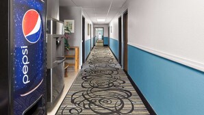 Corredor de hotel com uma máquina de refrigerantes Pepsi, carpete estampado e paredes azuis.