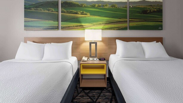 Quarto de hotel com duas camas, mesa de cabeceira com abajur, relógio e quadro de um campo de golfe.