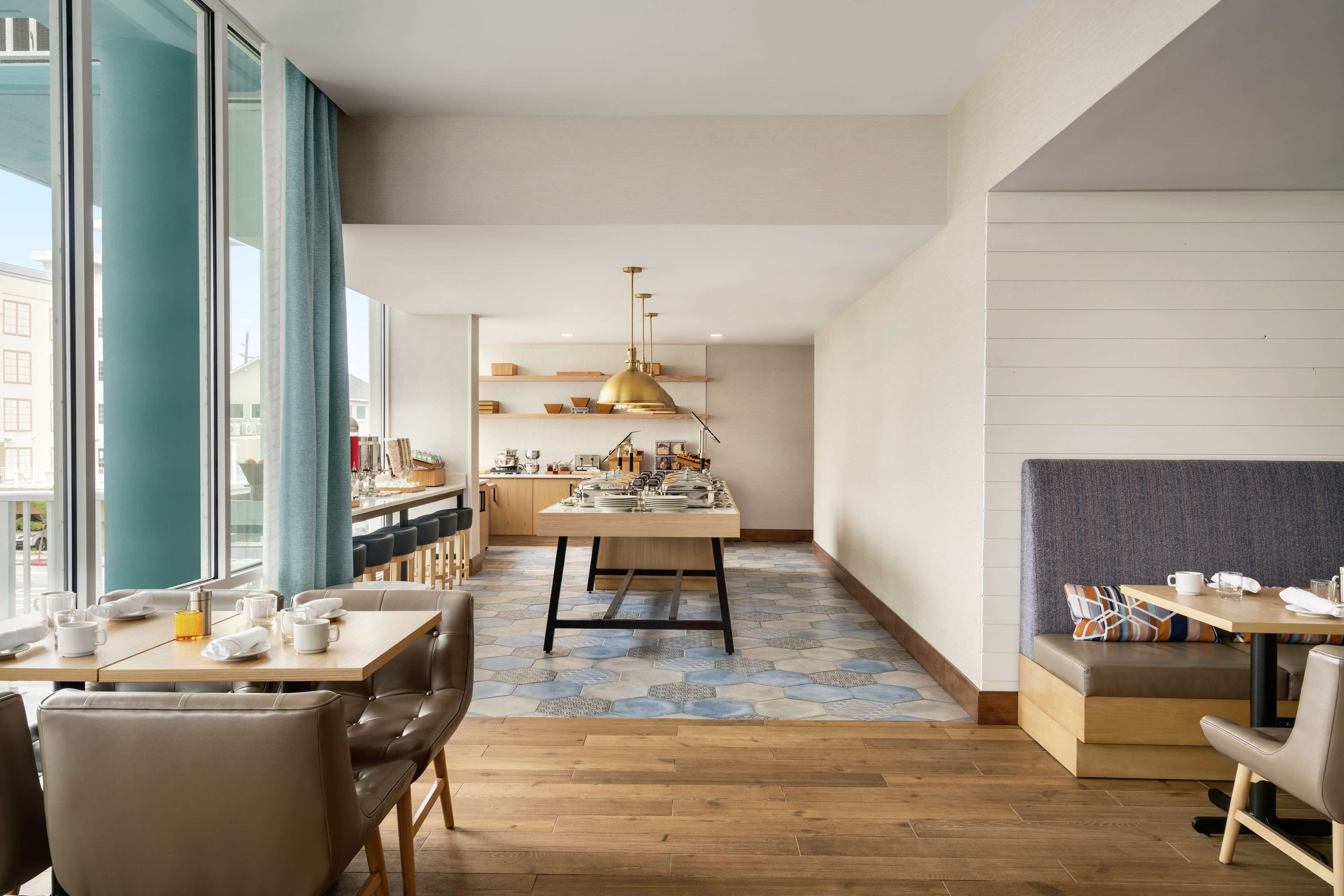 Uma zona de refeições moderna com soalho de madeira, uma mesa central com candeeiro pendente dourado e um arranjo de assentos com bancos estofados.
