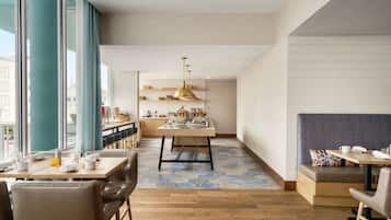 Uma zona de refeições moderna com soalho de madeira, uma mesa central com candeeiro pendente dourado e um arranjo de assentos com bancos estofados.