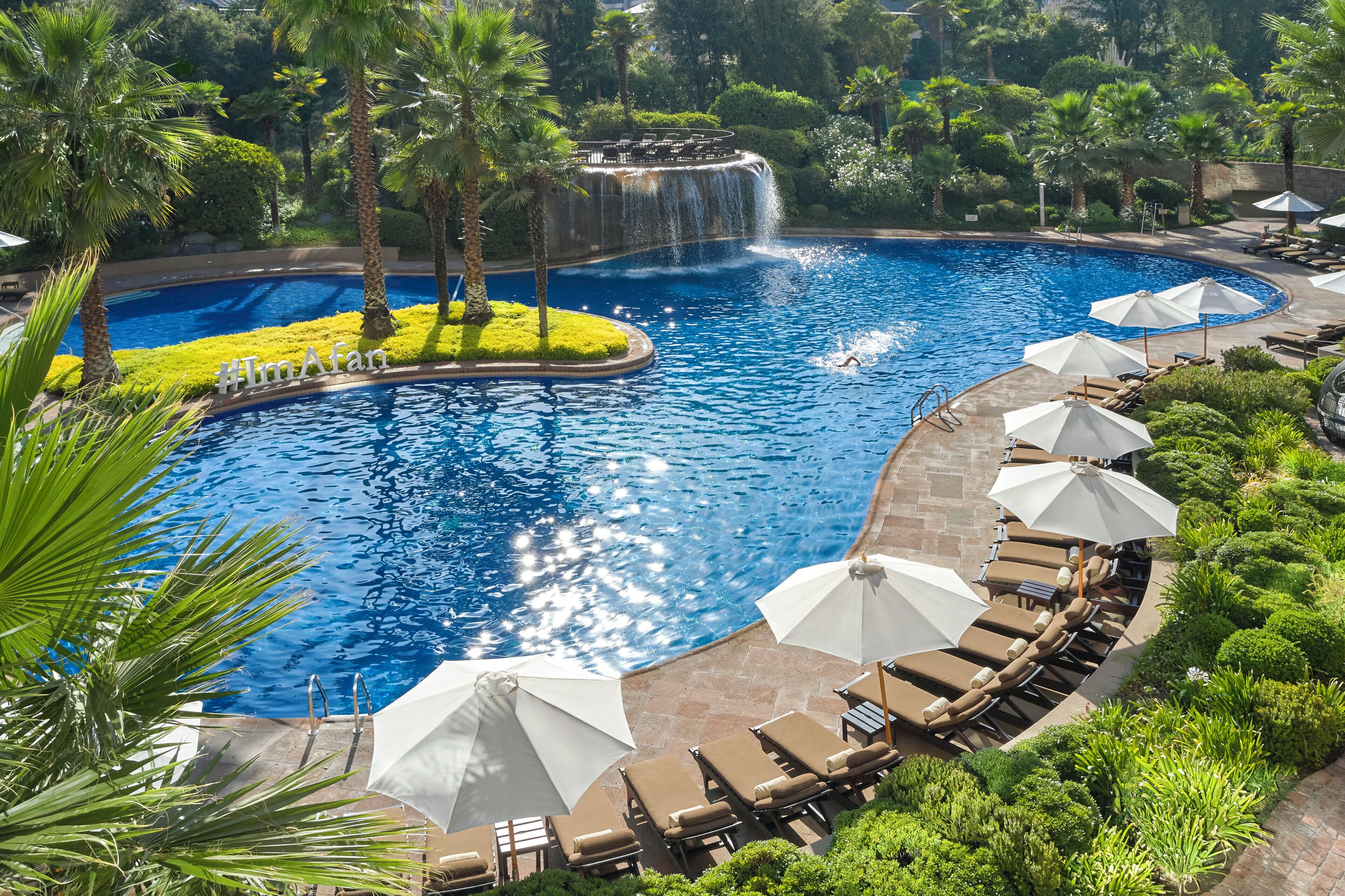 Piscina com cachoeira, cercada por palmeiras e espreguiçadeiras com guarda-sóis.