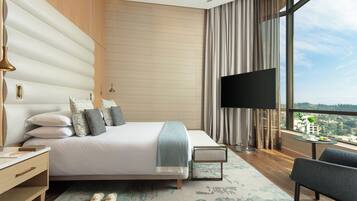 Quarto de hotel moderno com uma cama grande, televisão, vista da cidade e varanda com mesa e cadeira.